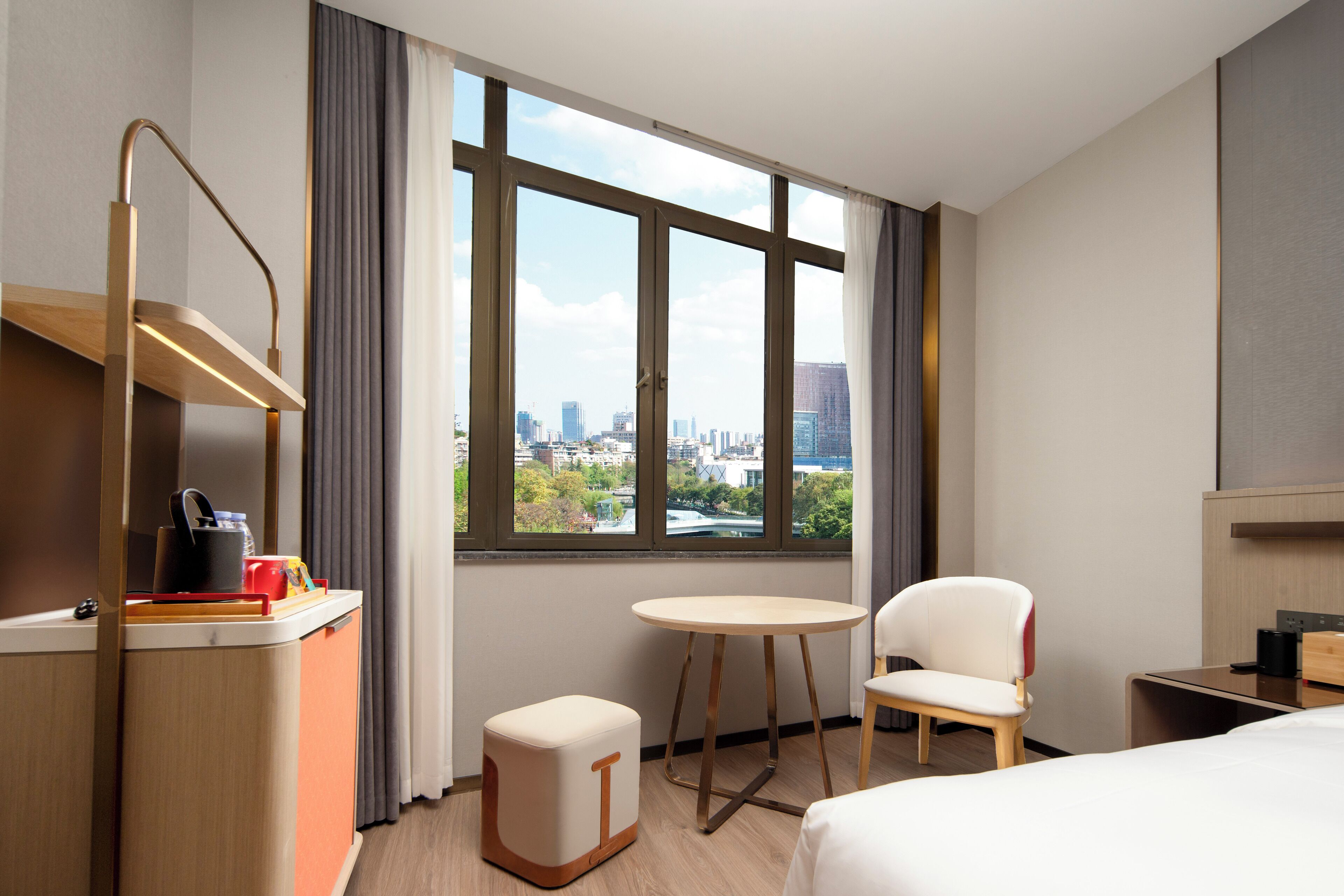 Quarto de hotel com uma cama, escrivaninha, cadeira, mesa pequena, abajur e janela com vista para a cidade.