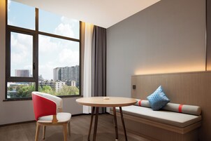 Quarto de hotel moderno com uma janela grande, uma mesa redonda, uma cadeira vermelha e branca e um sofá com almofada azul.
