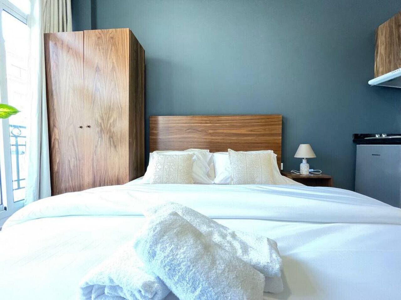Quarto de hotel com uma cama grande, guarda-roupa de madeira e uma pequena mesa de cabeceira com abajur.