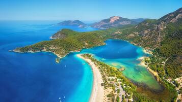 Vista aérea de uma área costeira com praia, águas azul-claras e colinas verdes ao redor.