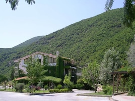 Um edifício de dois andares com um jardim na frente, cercado por vegetação exuberante e colinas.