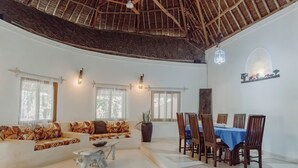 Uma sala de estar espaçosa com teto alto, uma mesa de jantar posta para seis pessoas e um sofá com almofadas tropicais.