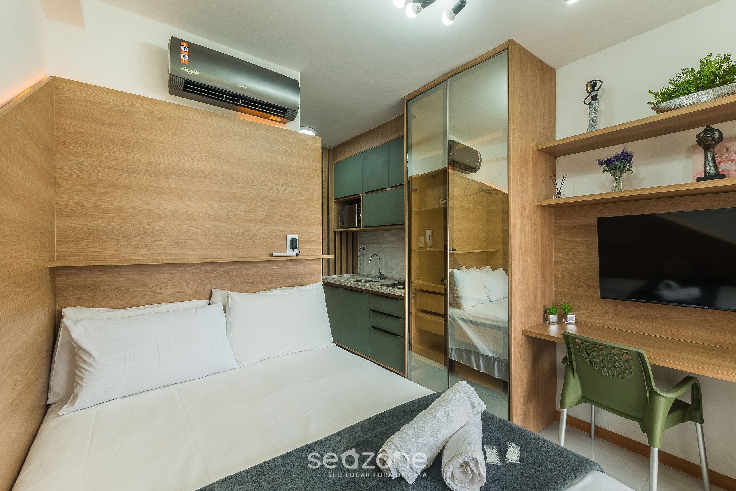 Quarto de hotel moderno com cama, secretária, cadeira, televisão e cozinha compacta.