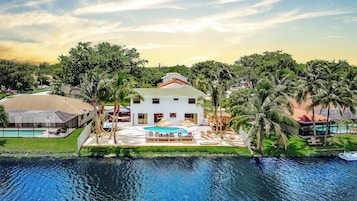 Uma casa grande e branca com piscina, cercada por palmeiras e um corpo de água.