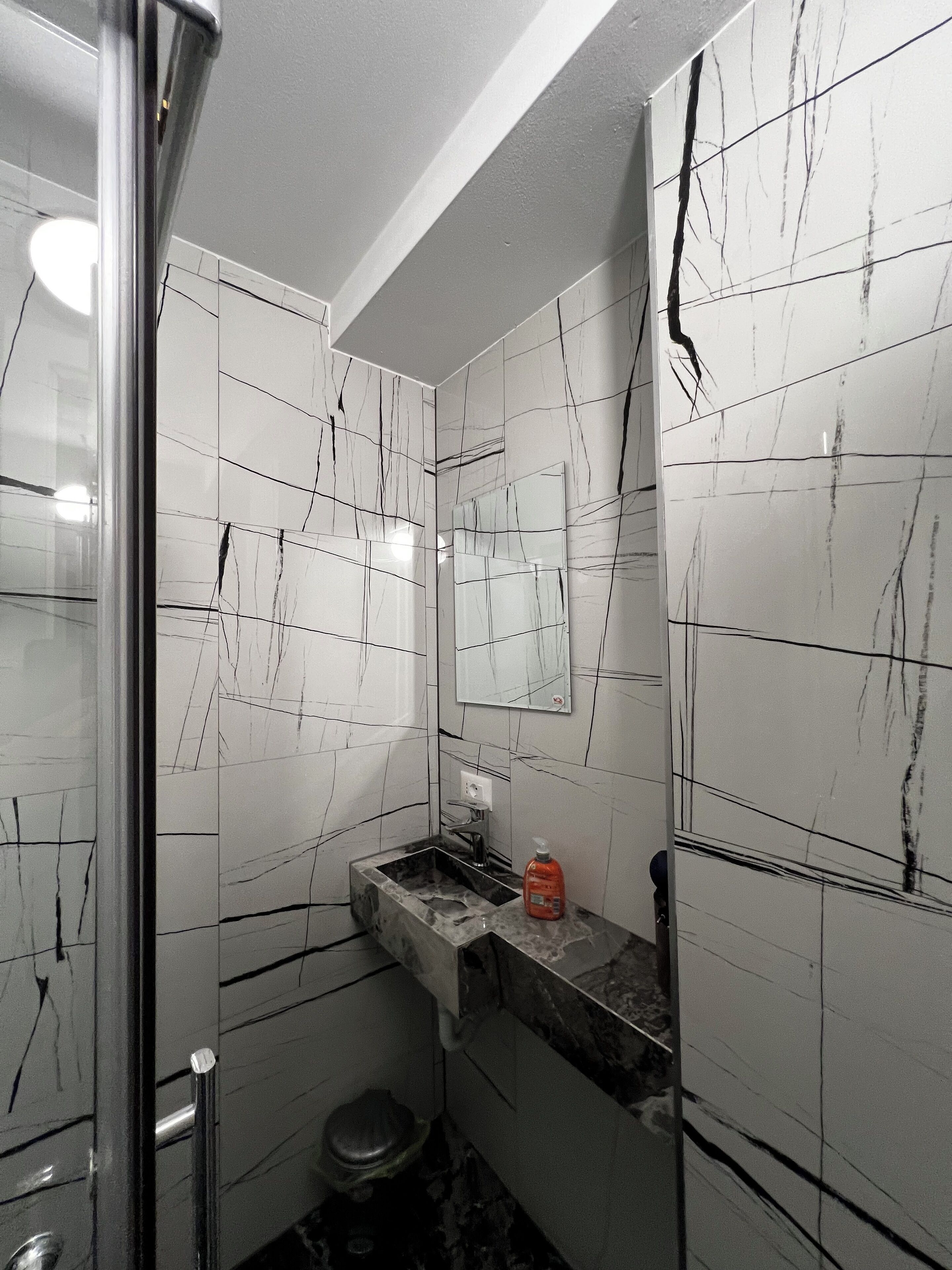 Um banheiro com azulejos de mármore branco, box de vidro, pia com espelho acima e vaso sanitário.