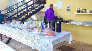 Disposição de buffet com uma mesa coberta por um mantel de renda, alimentos e um canto para café.
