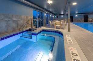 Piscina interna com marcações claras de profundidade, área de estar com mesas e cadeiras, e vista para o exterior através de amplas janelas.