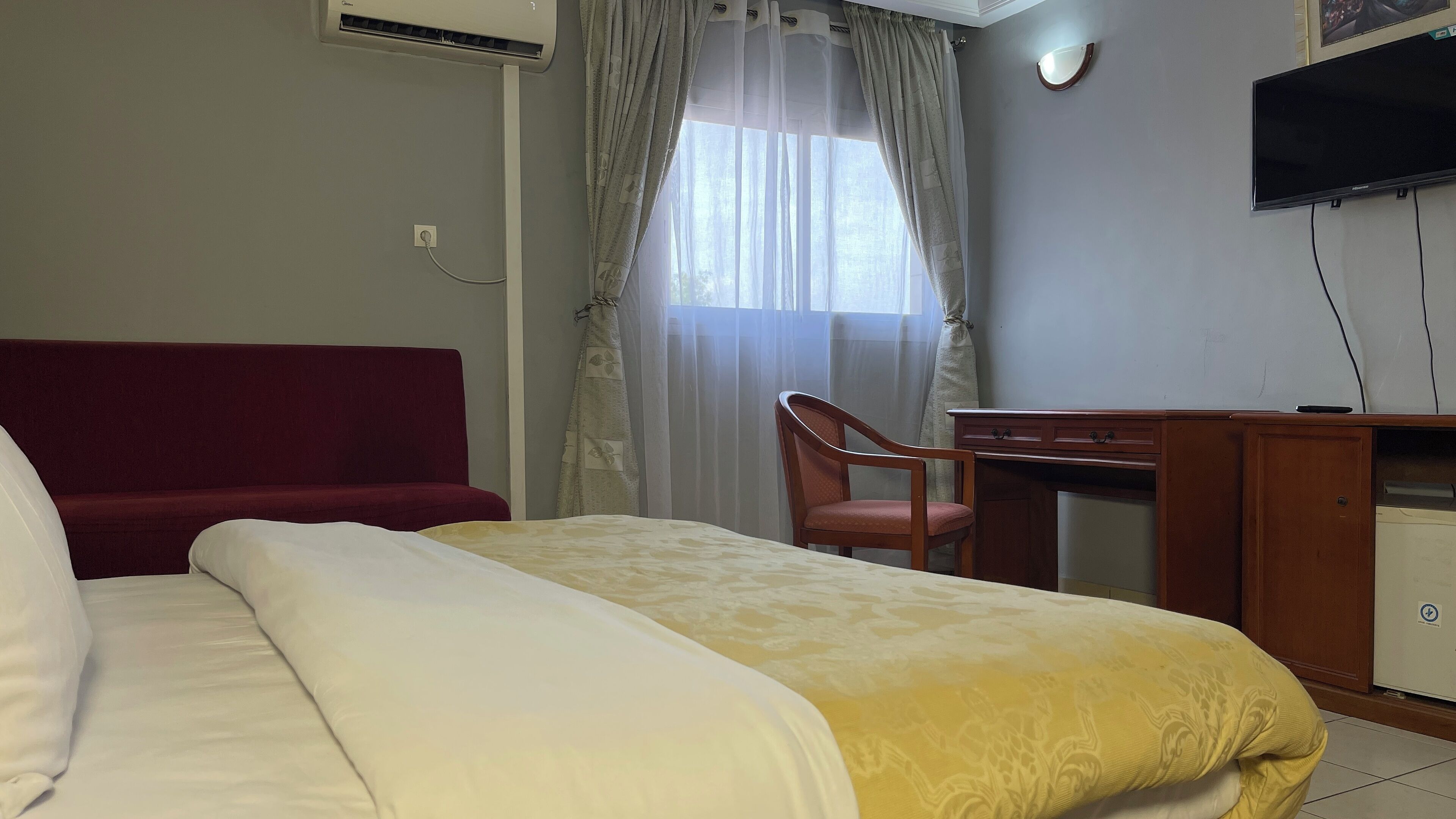 Quarto de hotel com cama, escrivaninha, cadeira, televisão e janela com cortinas.