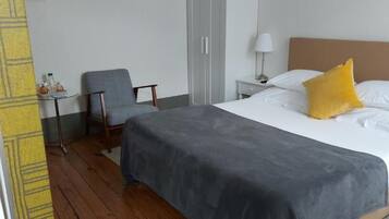 Quarto de hotel com cama, uma cadeira, uma mesinha com garrafas e um abajur.