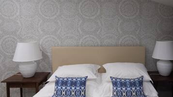 Cama bem arrumada com lençóis brancos e duas almofadas decorativas, com duas mesas de cabeceira com abajures, em frente a um papel de parede estampado.