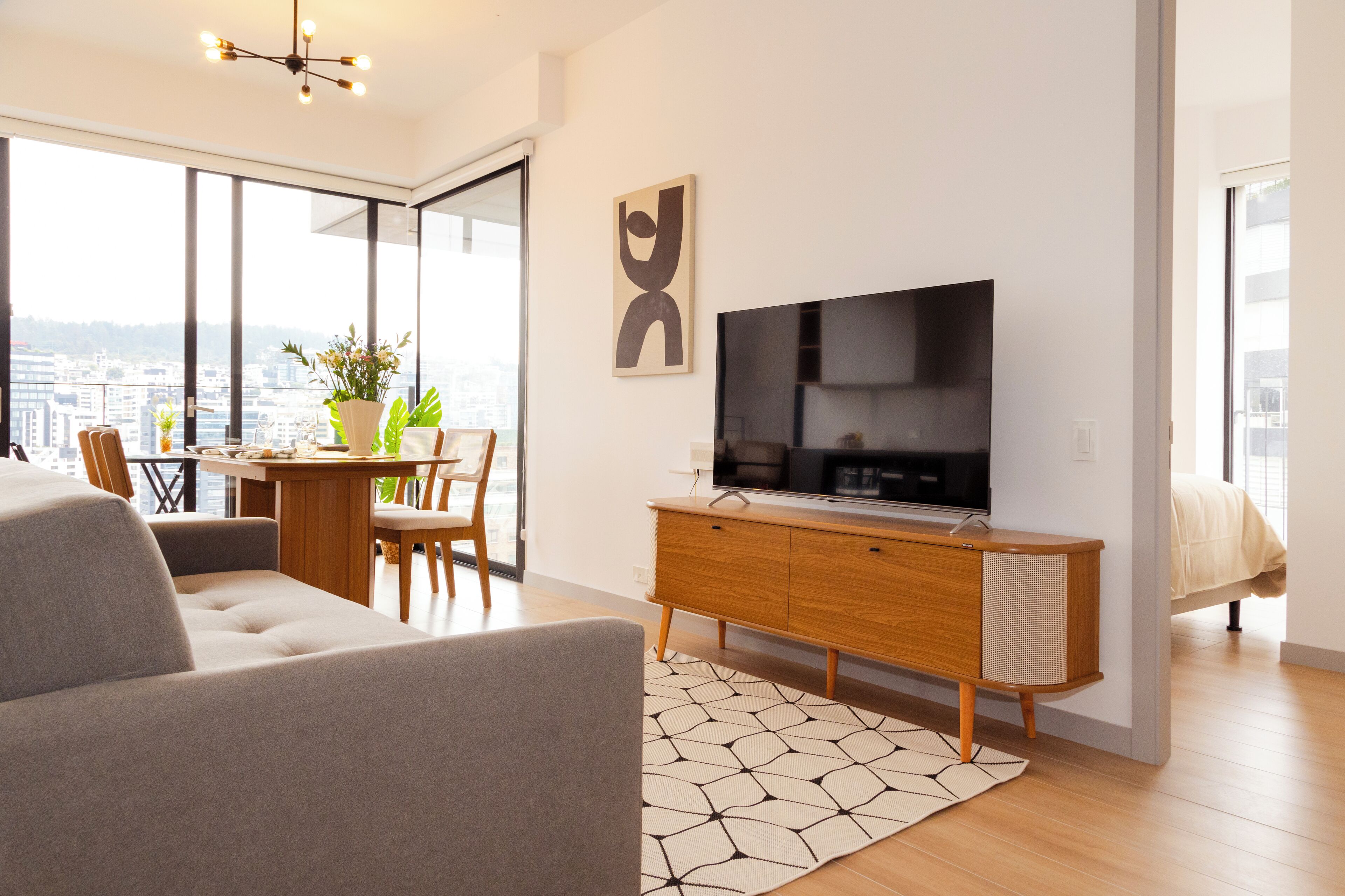 Uma sala de estar moderna com uma televisão grande, uma área de jantar com mesa e cadeiras, e vista da cidade através de grandes janelas.