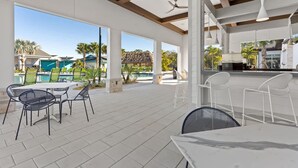 Uma área de terraço ao ar livre com mesas e cadeiras, uma piscina ao fundo e vista para outros edifícios.