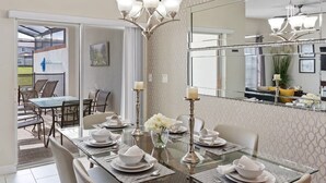 Uma sala de jantar com mesa de vidro posta para seis, um candelabro e um espelho grande refletindo o ambiente.