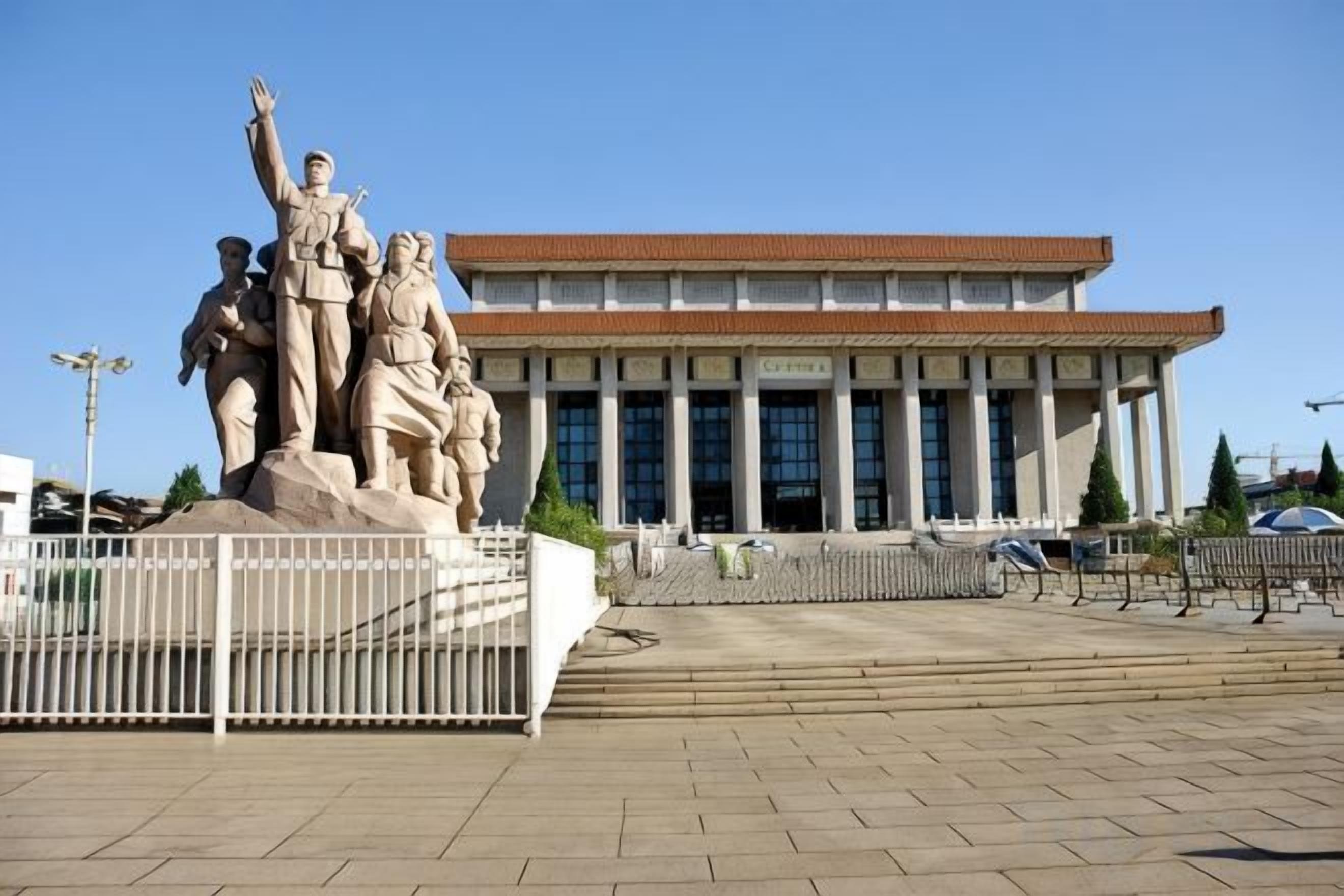 Um edifício com estilo arquitetônico clássico, apresentando uma estátua central em pedestal, janelas amplas e uma área de entrada pavimentada.