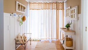 Quarto com um banco, uma pequena mesa com uma planta, um cesto e uma janela com cortinas.