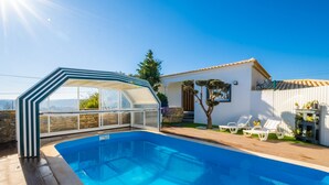 Uma piscina com cobertura de vidro, cadeiras de lounge brancas e uma pequena casa com telhado em azulejos.