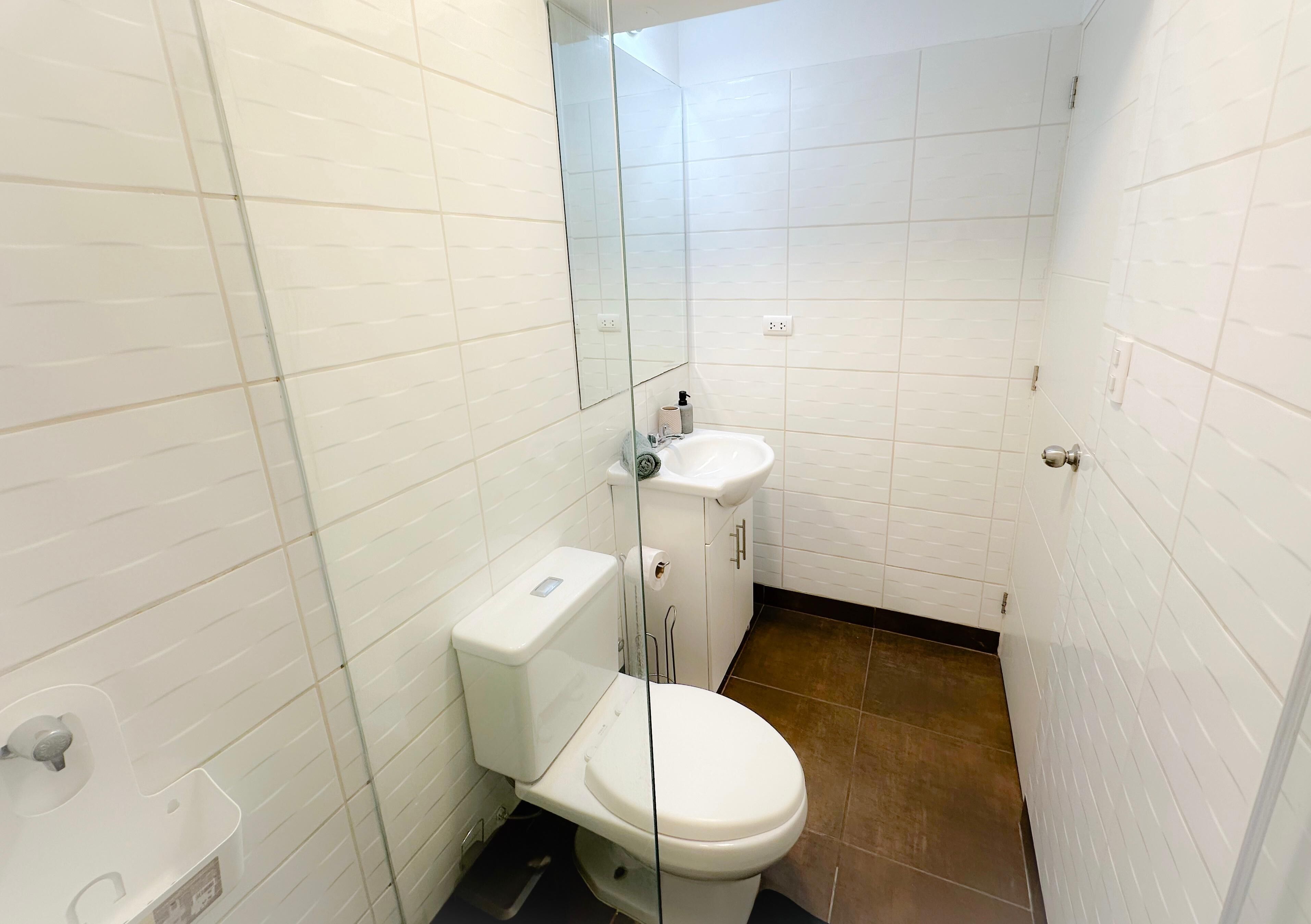 Uma casa de banho pequena e limpa, com azulejos brancos, sanita, lavatório e espelho.