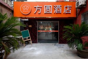 Entrada de hotel com placa laranja chamativa, porta de vidro e plantas em vasos.