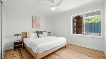 Quarto com cama de madeira, paredes brancas, ventoinha no teto, janela com vista para uma parede de tijolos e vegetação, e mesa de cabeceira com abajur.