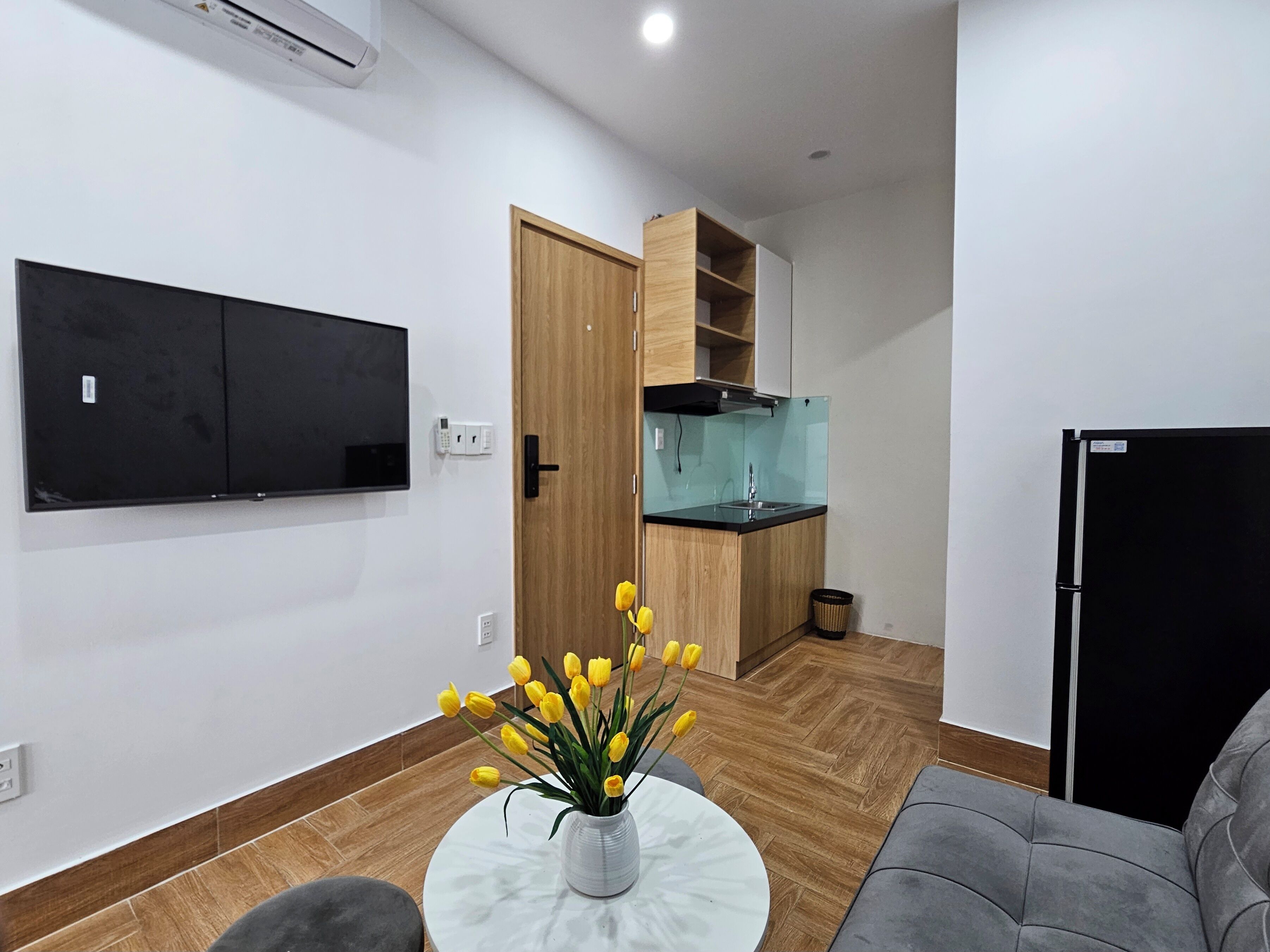 Uma sala de estar moderna com um sofá cinzento, uma mesa de centro branca com um vaso de flores amarelas, uma televisão de tela plana na parede e uma geladeira preta.