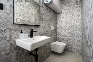 Casa de banho com lavatório branco, torneira preta e sanita. As paredes apresentam um padrão geométrico em azulejos.