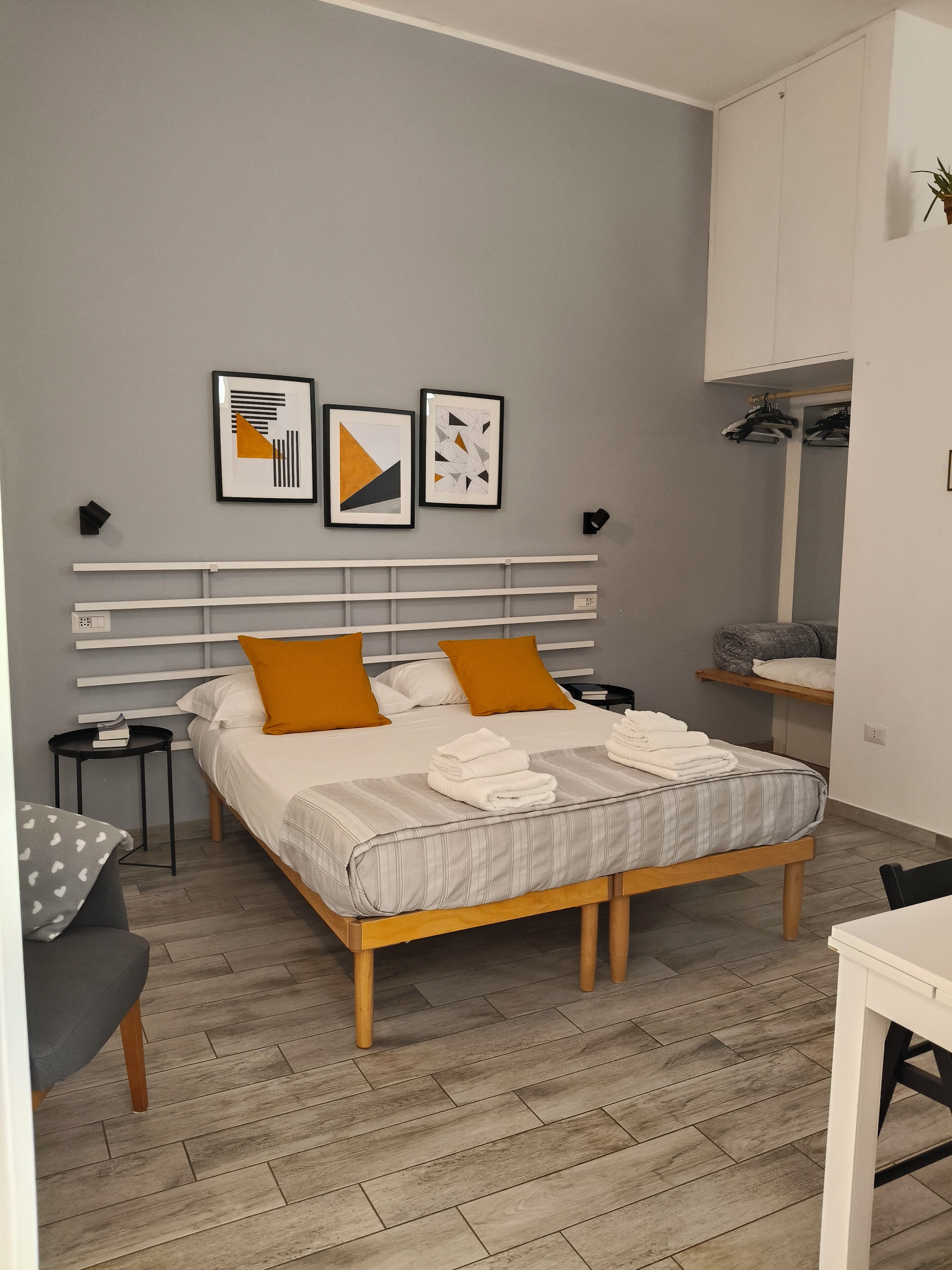 Quarto moderno com cama, duas almofadas, mesa de cabeceira, cadeira, escrivaninha, guarda-roupa e três obras de arte emolduradas na parede.