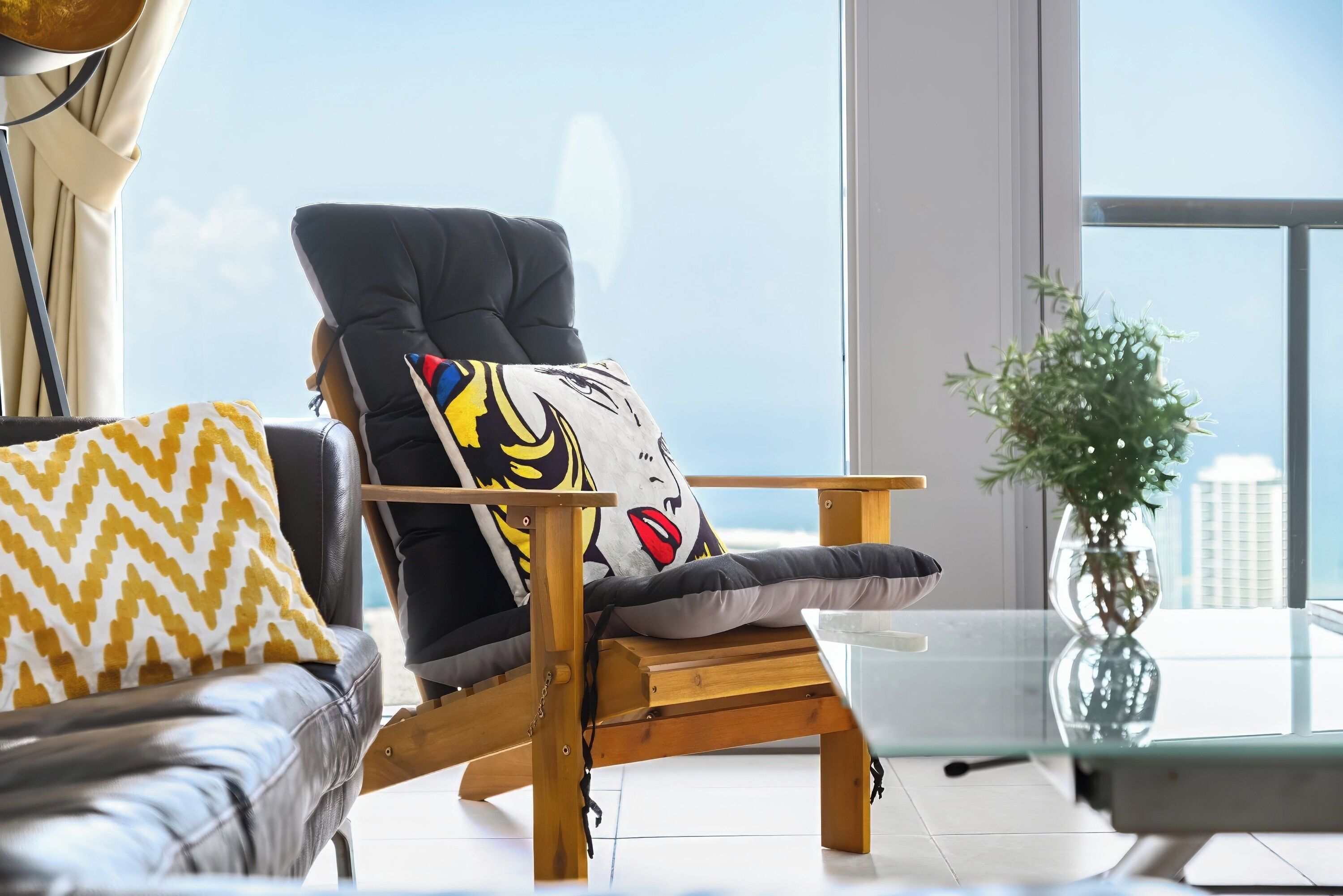 Uma sala de estar moderna com um sofá de couro preto, uma cadeira de madeira com almofada colorida e uma mesa de centro de vidro.