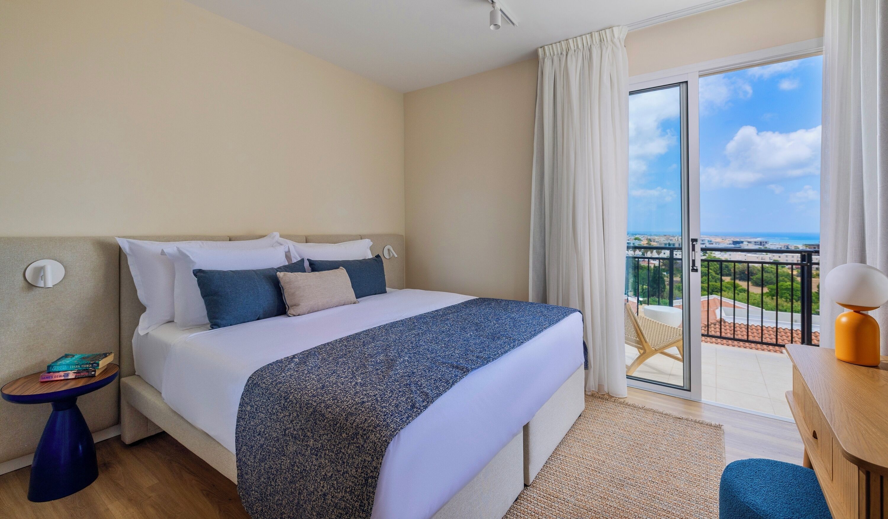 Quarto de hotel com uma cama, uma pequena mesa com livros, uma cadeira e vista para o mar através de uma varanda.