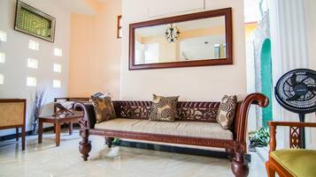 Uma sala de estar com sofá de madeira, mesa de jantar, cadeiras, um espelho grande e um ventilador de pé.