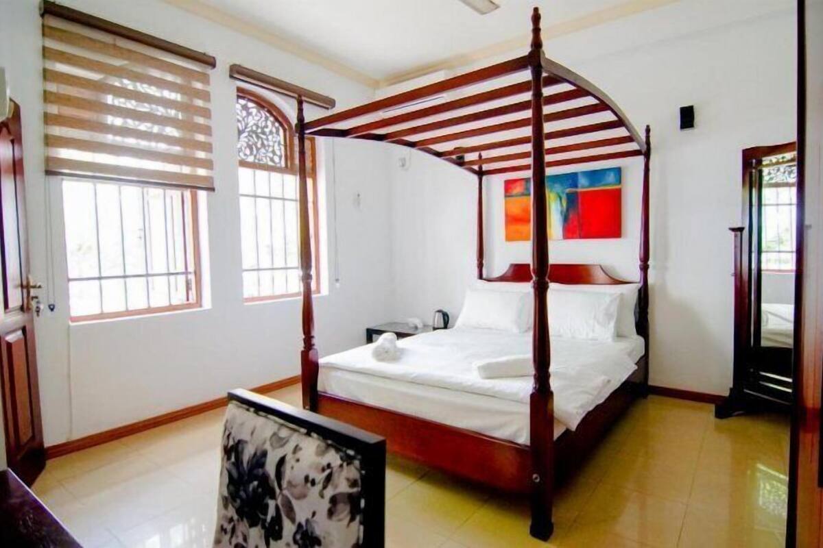 Uma cama com dossel, colchão branco, chão de madeira e um quadro na parede.