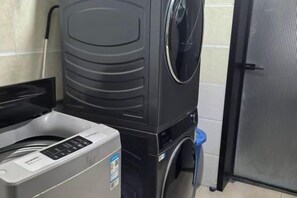 Uma lavanderia compacta e moderna, com máquina de lavar e secadora frontais, uma pequena lixeira e uma porta que dá para outro cômodo.