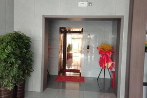 Entrada com tapete vermelho, planta em vaso e arranjo floral vermelho e amarelo.