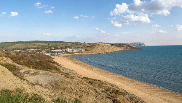 Paisagem costeira com uma praia arenosa, céu azul límpido e uma vista distante de uma cidade com edifícios.