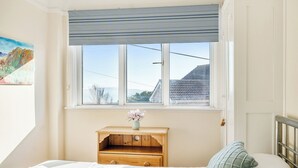Quarto com uma cama, uma mesinha de cabeceira de madeira com um vaso de flores, uma janela com persiana listrada e vista para uma casa lá fora.