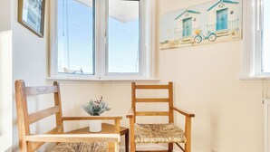 Quarto com duas cadeiras de madeira, uma mesa pequena e janela com vista para casas.