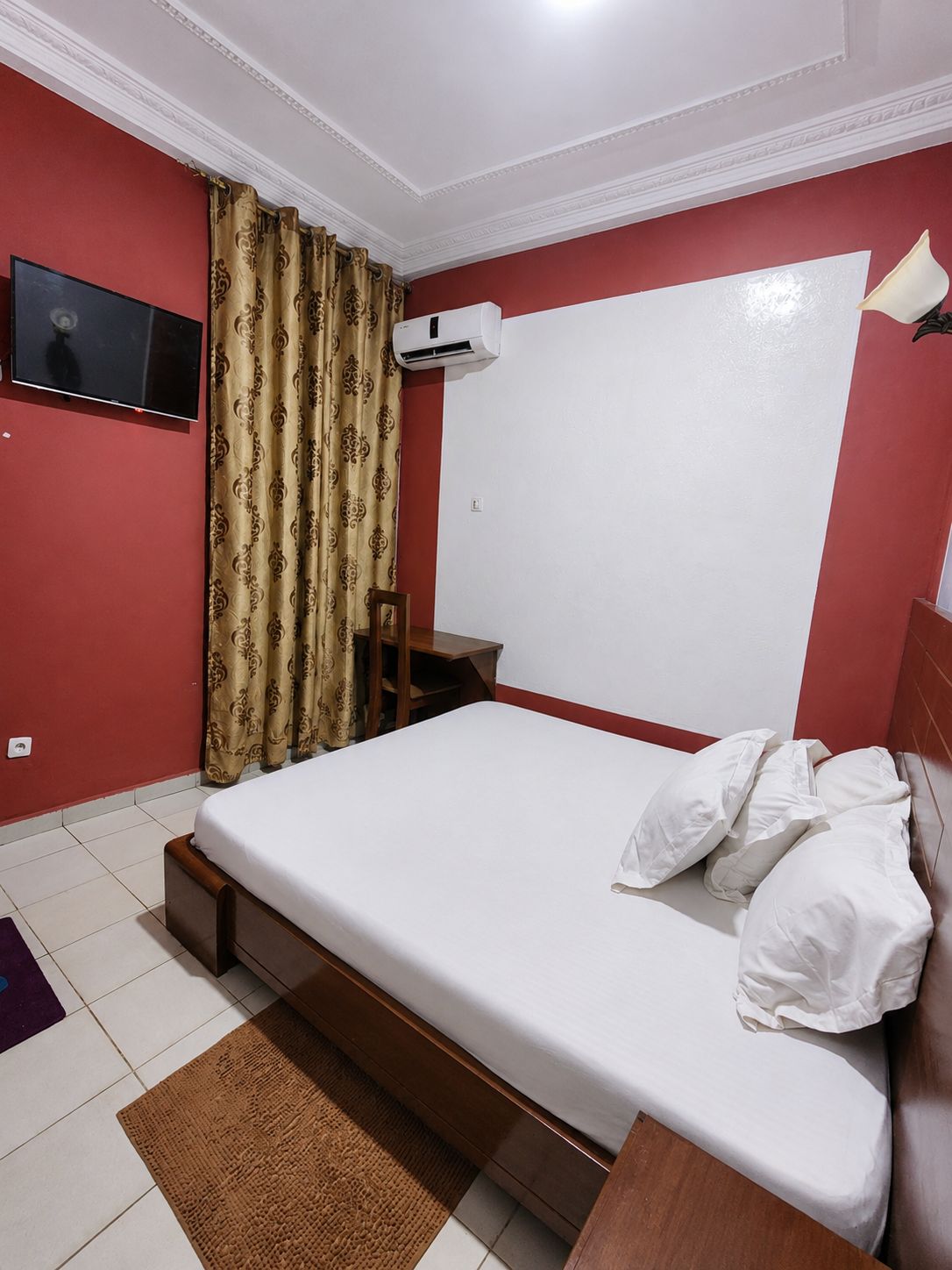 Quarto de hotel com cama, televisão de tela plana, ar condicionado, mesa de cabeceira de madeira e parede vermelha.
