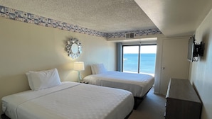 Quarto de hotel com duas camas, uma televisão na parede e vista para o mar através de uma grande janela.