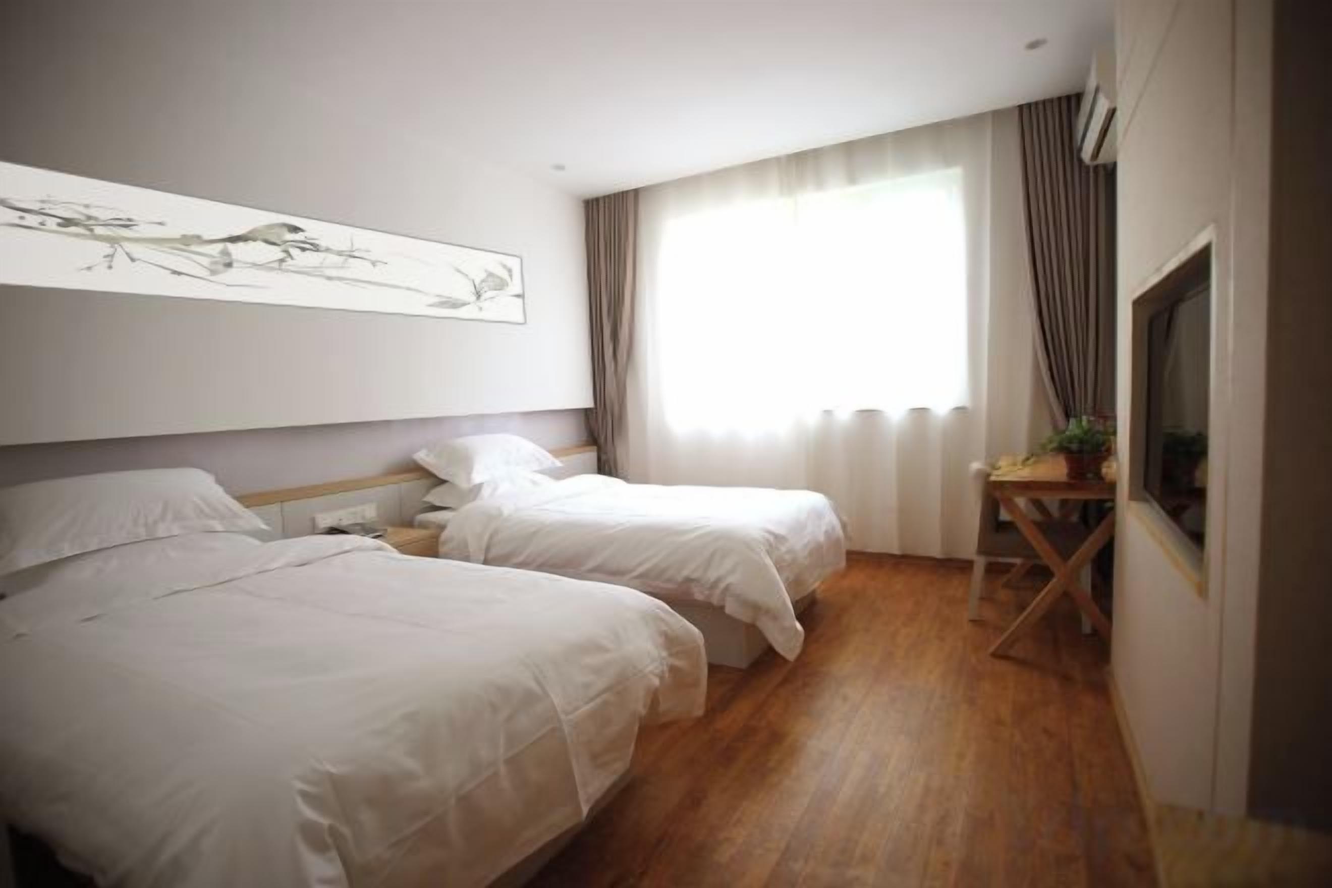 Quarto de hotel com duas camas, uma janela com cortinas, uma mesinha com uma planta e uma televisão fixada na parede.