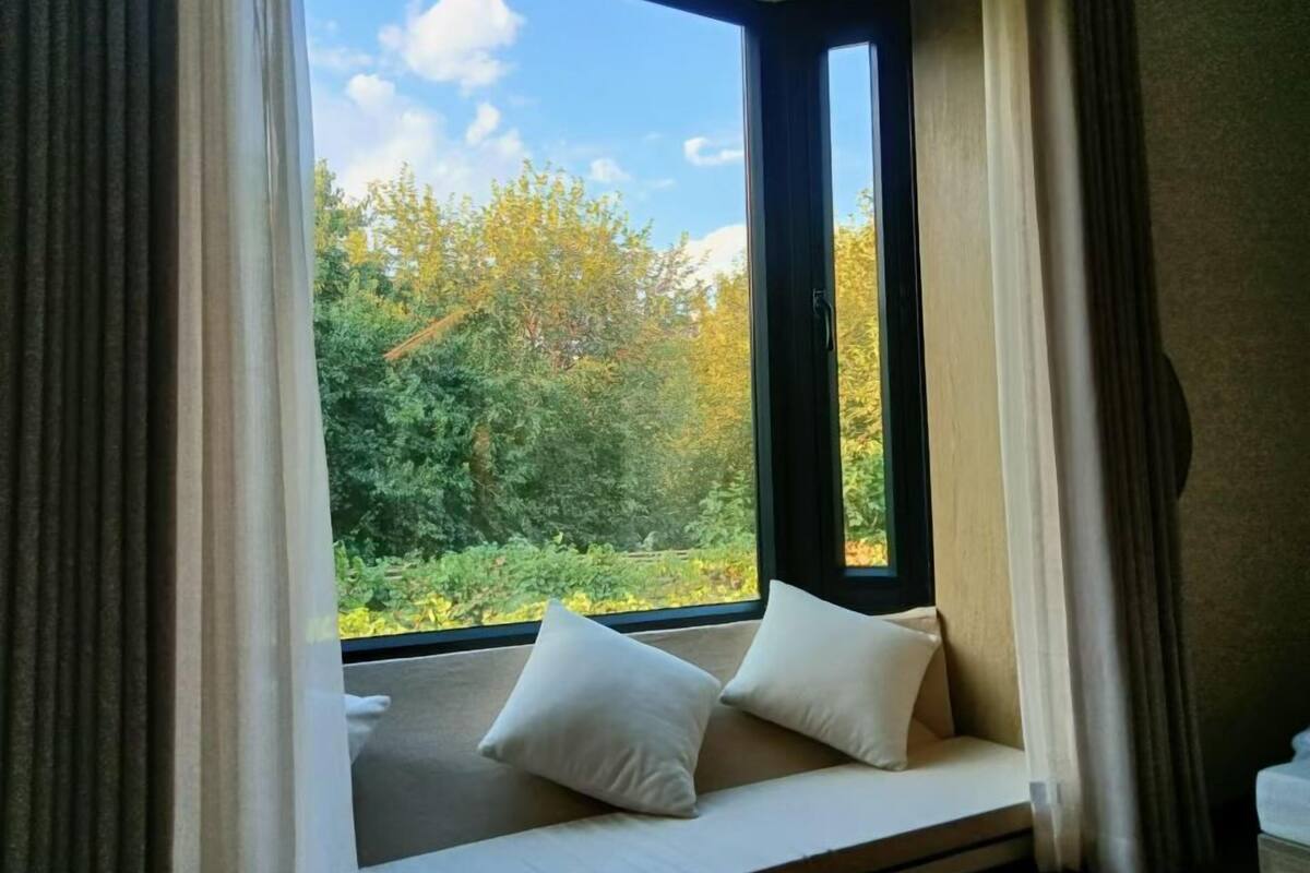 Quarto com um assento junto à janela, cortinas e vista para árvores.