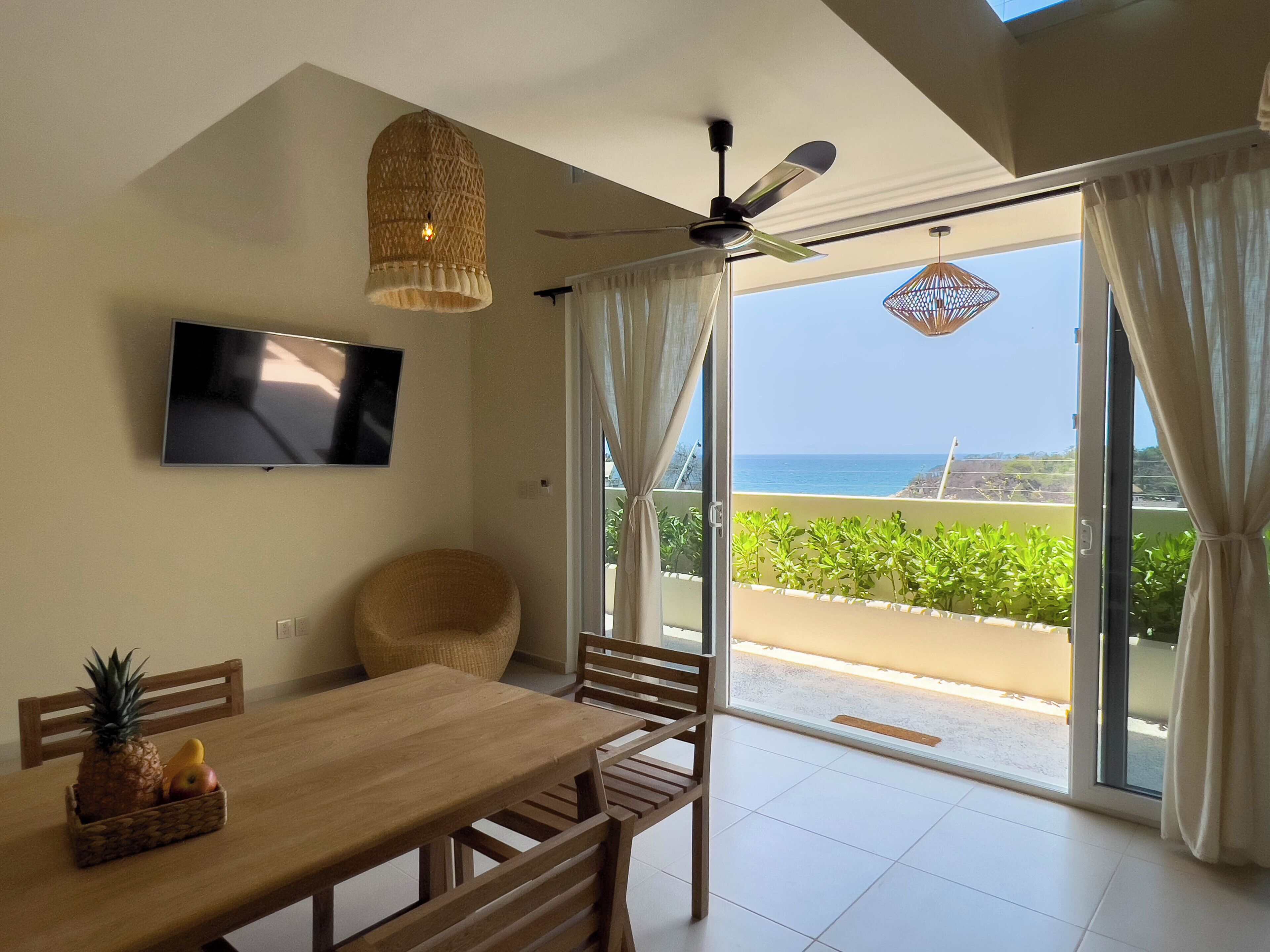 Uma sala de jantar com mesa, cadeiras, televisão e vista para o mar através de grandes janelas.