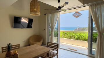 Uma sala de jantar com mesa, cadeiras, televisão e vista para o mar através de grandes janelas.