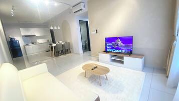 Uma sala de estar moderna com uma televisão de tela plana, um sofá branco, uma mesa de centro e uma cozinha ao fundo.