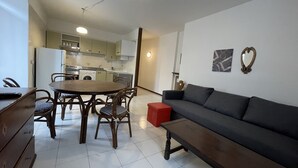 Uma sala moderna com um sofá, uma mesa de jantar com cadeiras, uma área de cozinha ao fundo e um banquinho vermelho.