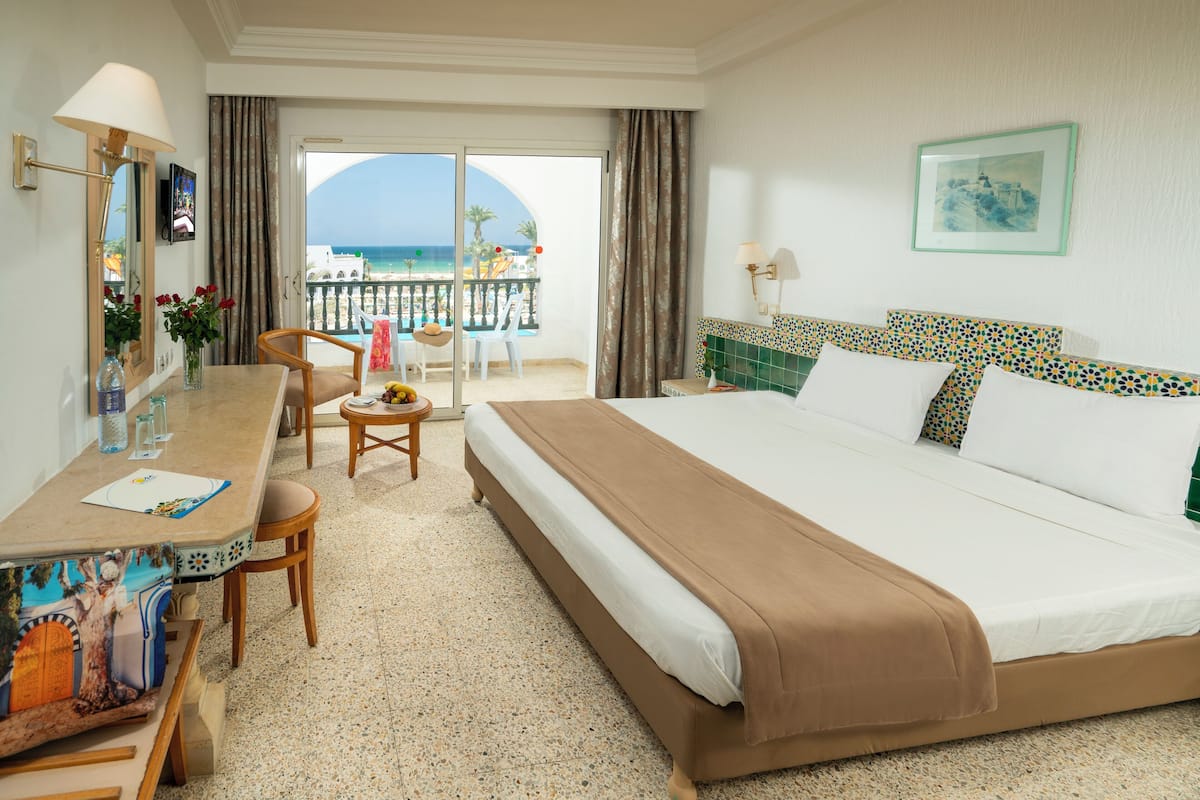 Quarto de hotel com uma cama grande, uma escrivaninha com cadeira, uma mesinha com cadeira, uma varanda com vista para a praia e um quadro na parede.