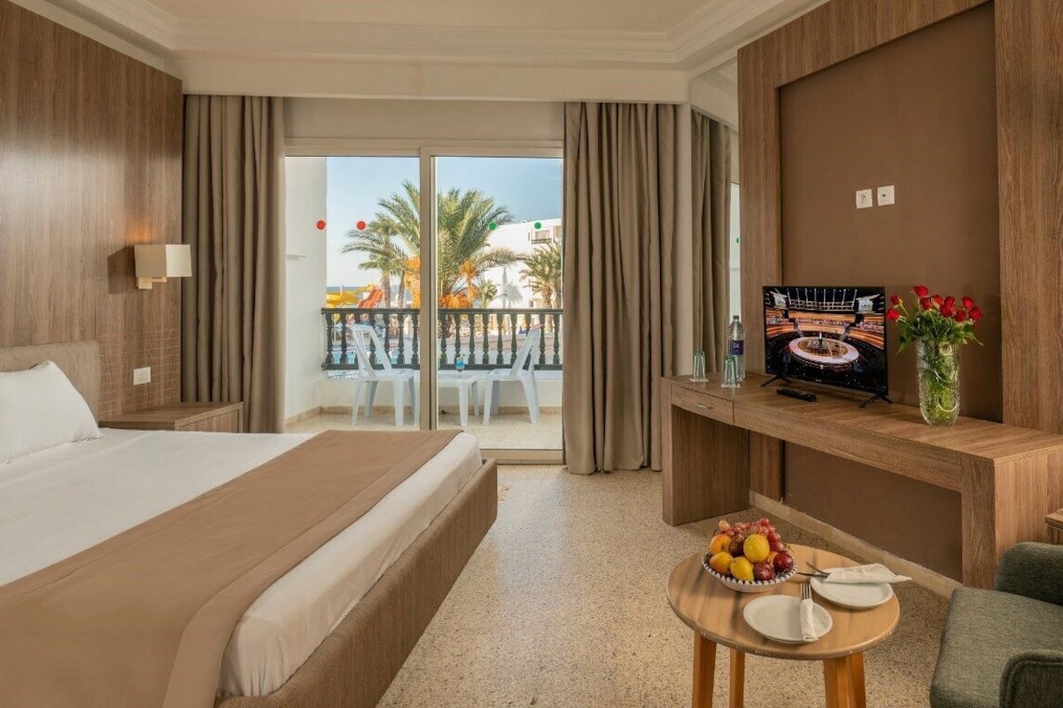 Quarto de hotel com uma cama grande, uma mesinha com frutas, televisão e varanda com área de estar ao ar livre.