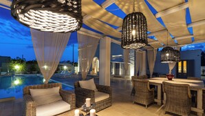 Uma área de estar externa com mobiliário de vime, piscina e luminárias pendentes.
