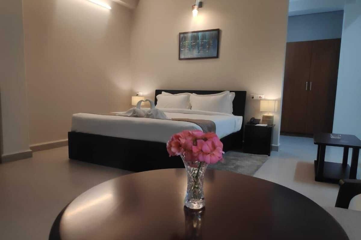Quarto de hotel com uma cama, mesas de cabeceira, uma mesa redonda com um vaso de flores e um quadro na parede.