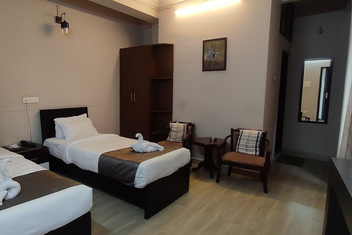 Quarto de hotel com duas camas, uma mesinha, uma cadeira, um guarda-roupa e um espelho.