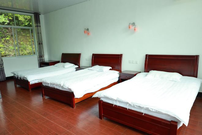 Quarto de hotel com três camas de madeira, uma janela grande com cortinas e luminárias fixadas na parede.