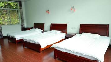 Quarto de hotel com três camas de madeira, uma janela grande com cortinas e luminárias fixadas na parede.